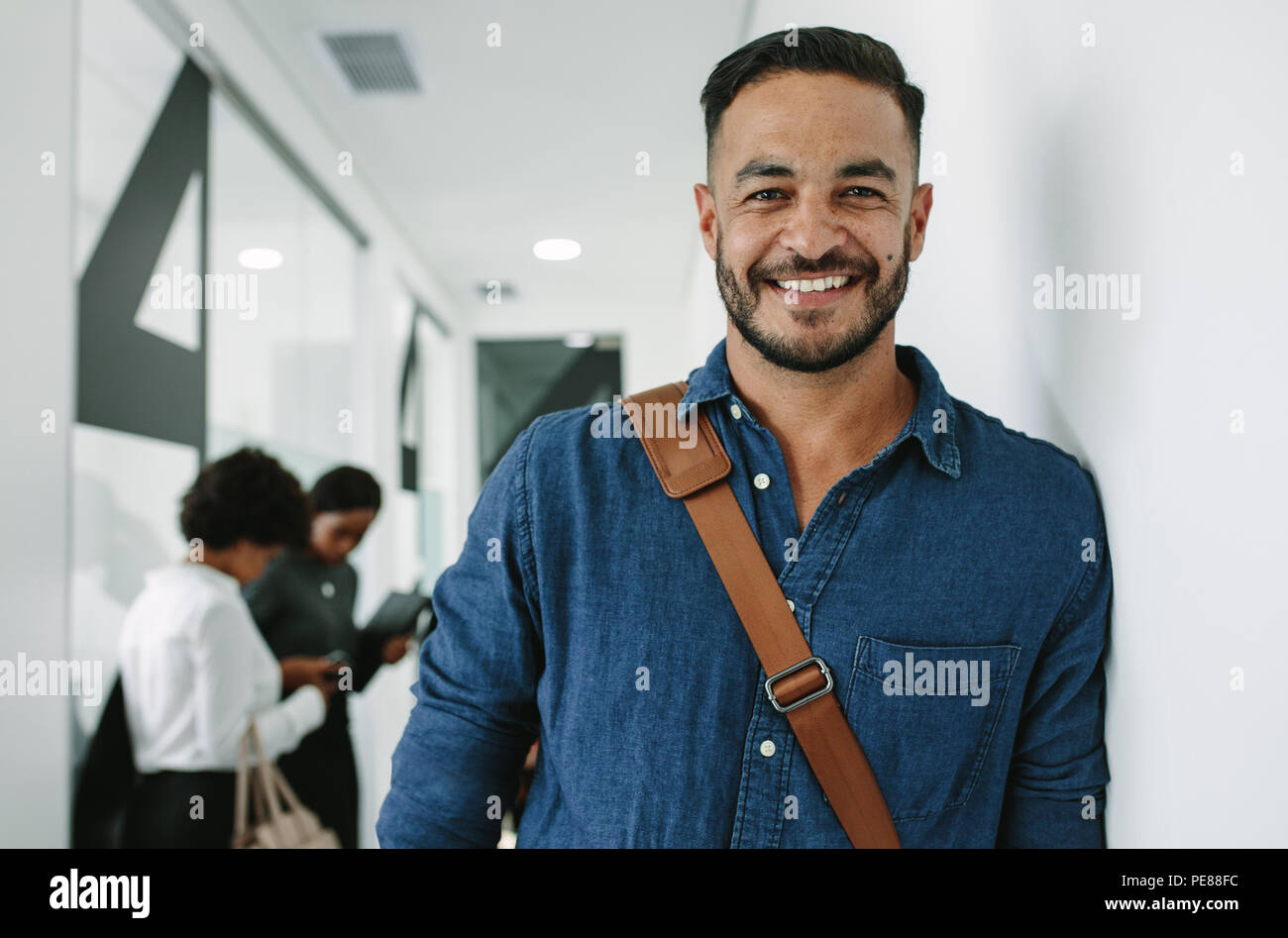 Portrait of young man in casuals standing at office corridor with ...