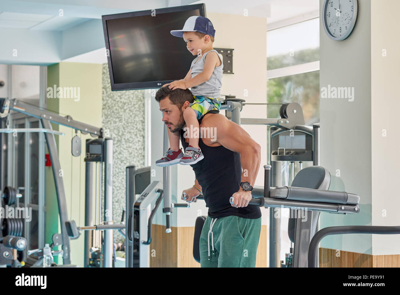 Fit sportsman training on gym's simulator keeping little son on neck ...
