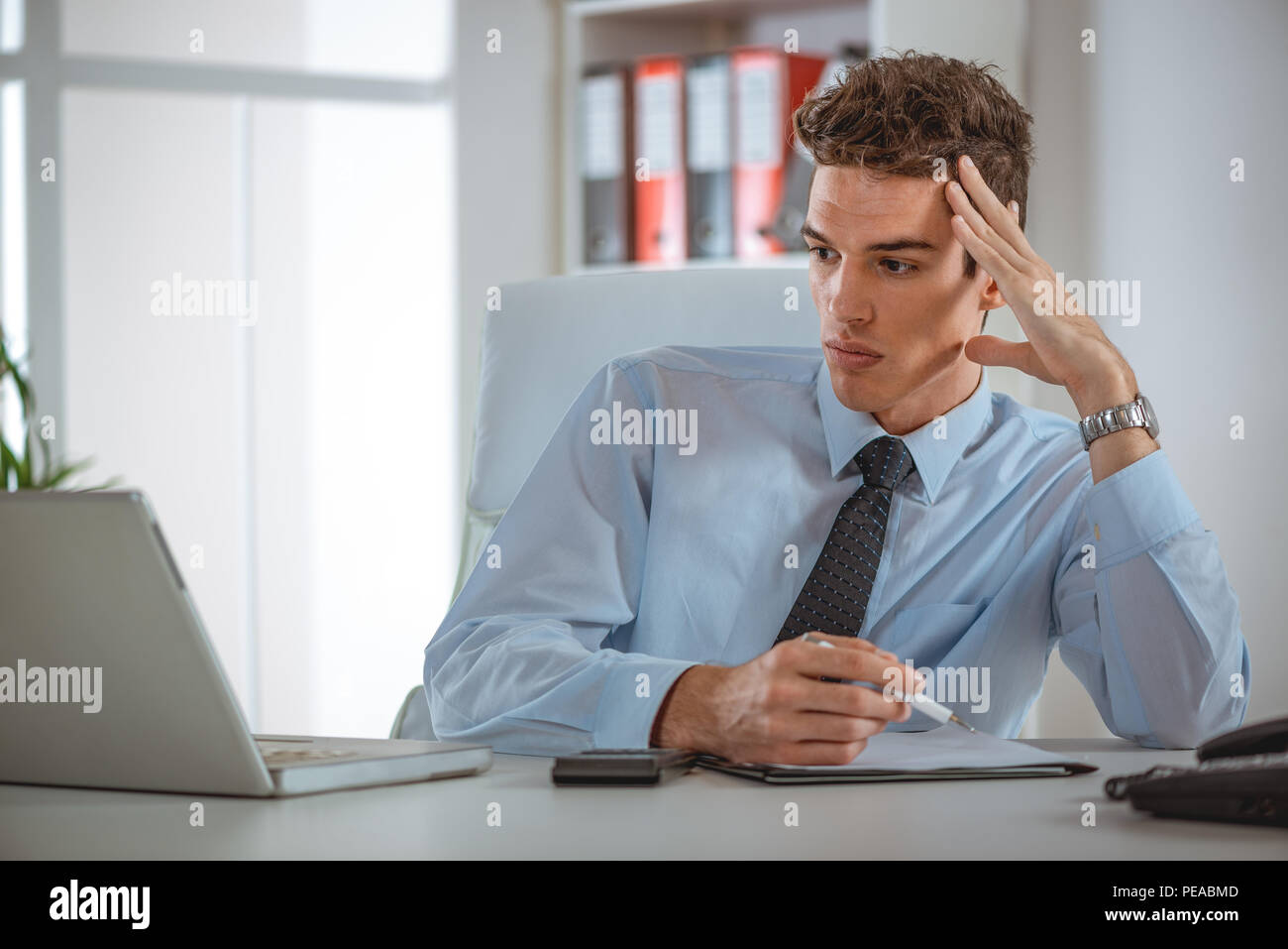 A serious young employee working in office with laptop and making notes ...