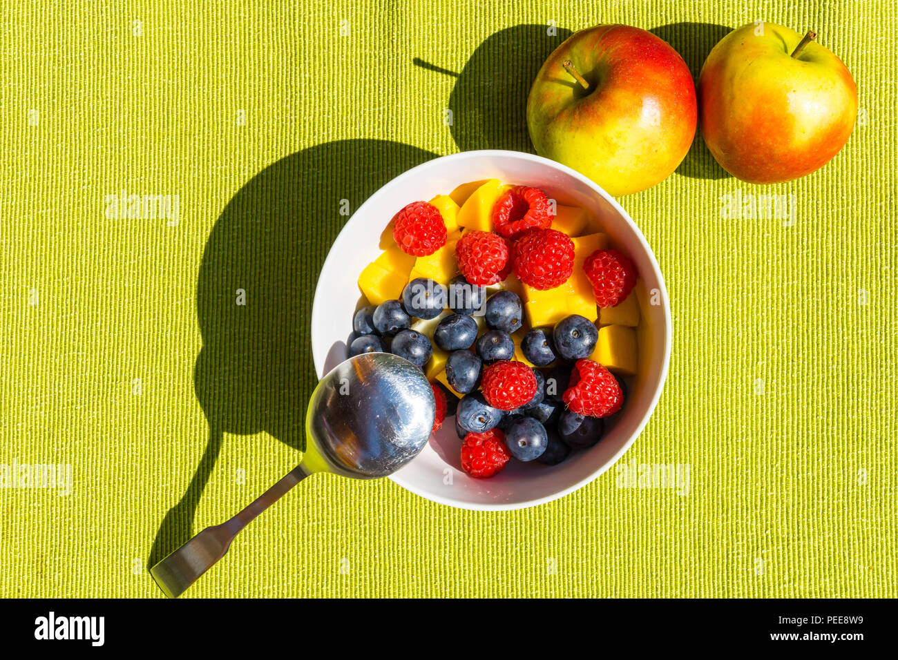 Mixed fruits of different color in a dish with two apples and a spoon ...