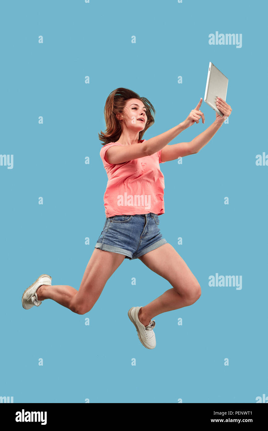 Jump of young woman over blue studio background using laptop or tablet ...