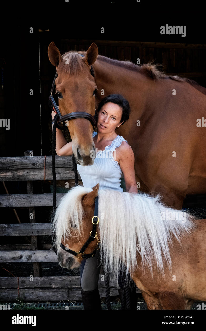 Woman standing outside a stable, holding a brown bay horse and a small ...