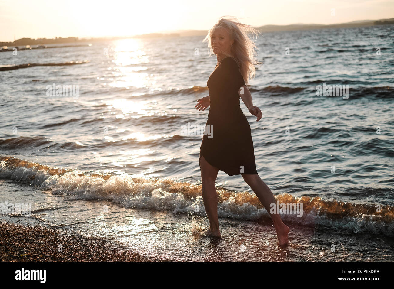Young blonde woman in black dress running by the sea, sunset light ...
