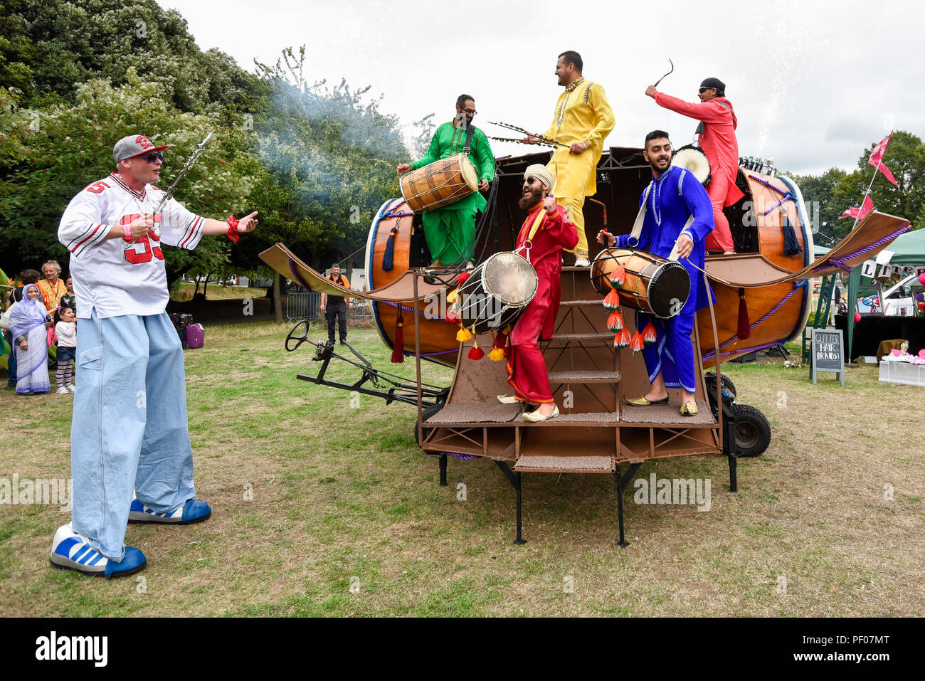 London, UK. 18 August 2018. A performer on stilts stands with drummers ...