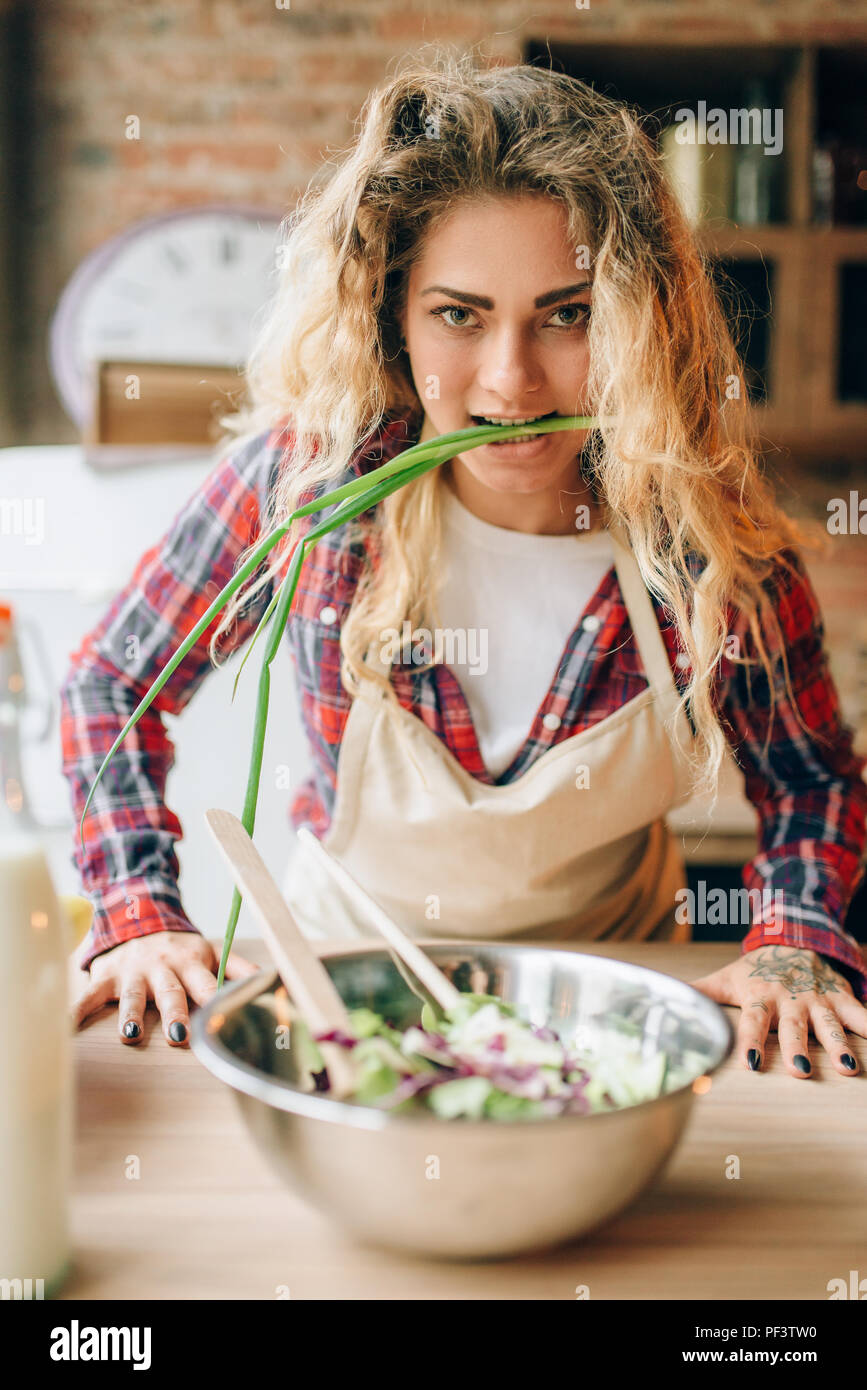 Housewife in an apron holds in teeth a brush of green onions, kitchen ...