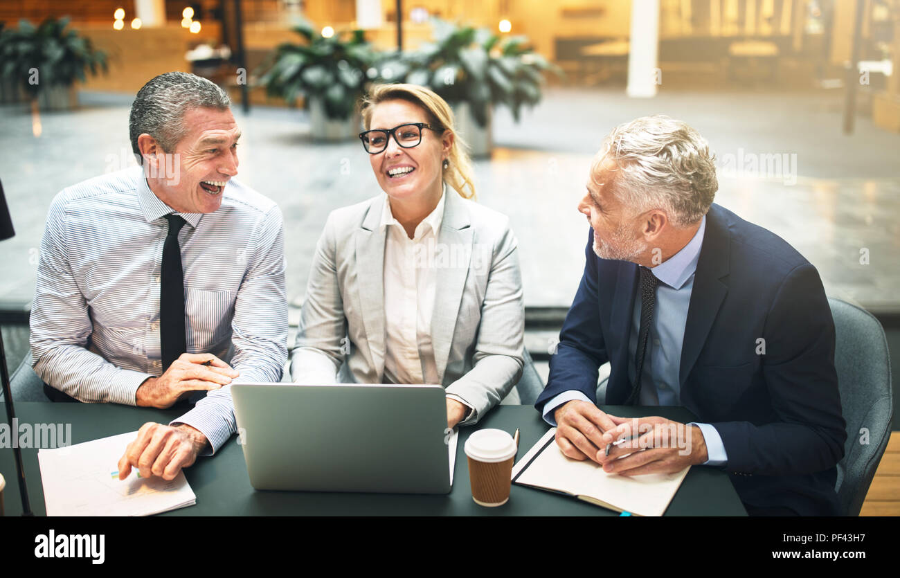 Three corporate colleagues laughing and talking together over a laptop at a table in the lobby ...