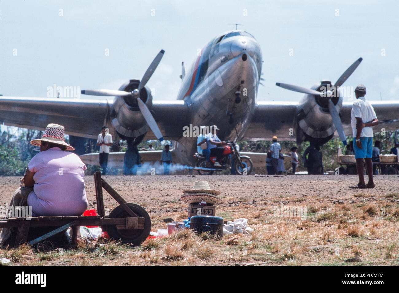 A vintage propeller plane at a rural airstrip, with locals gathered and ...