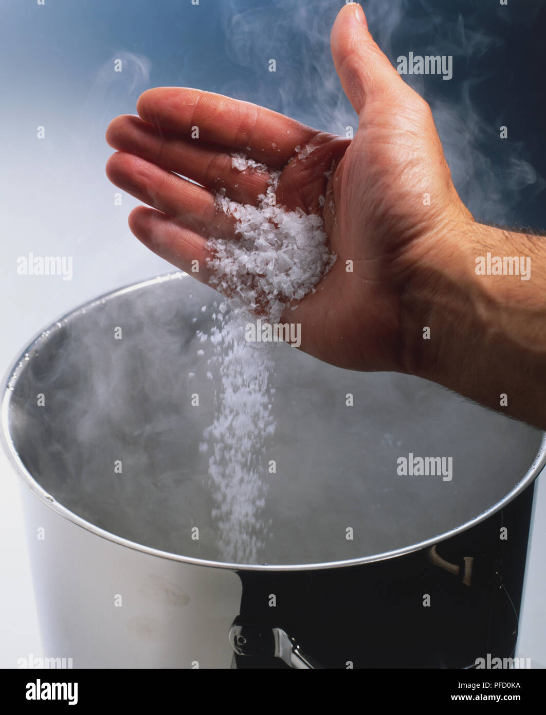 Hand pouring coarse salt grains into steaming saucepan, elevated view ...