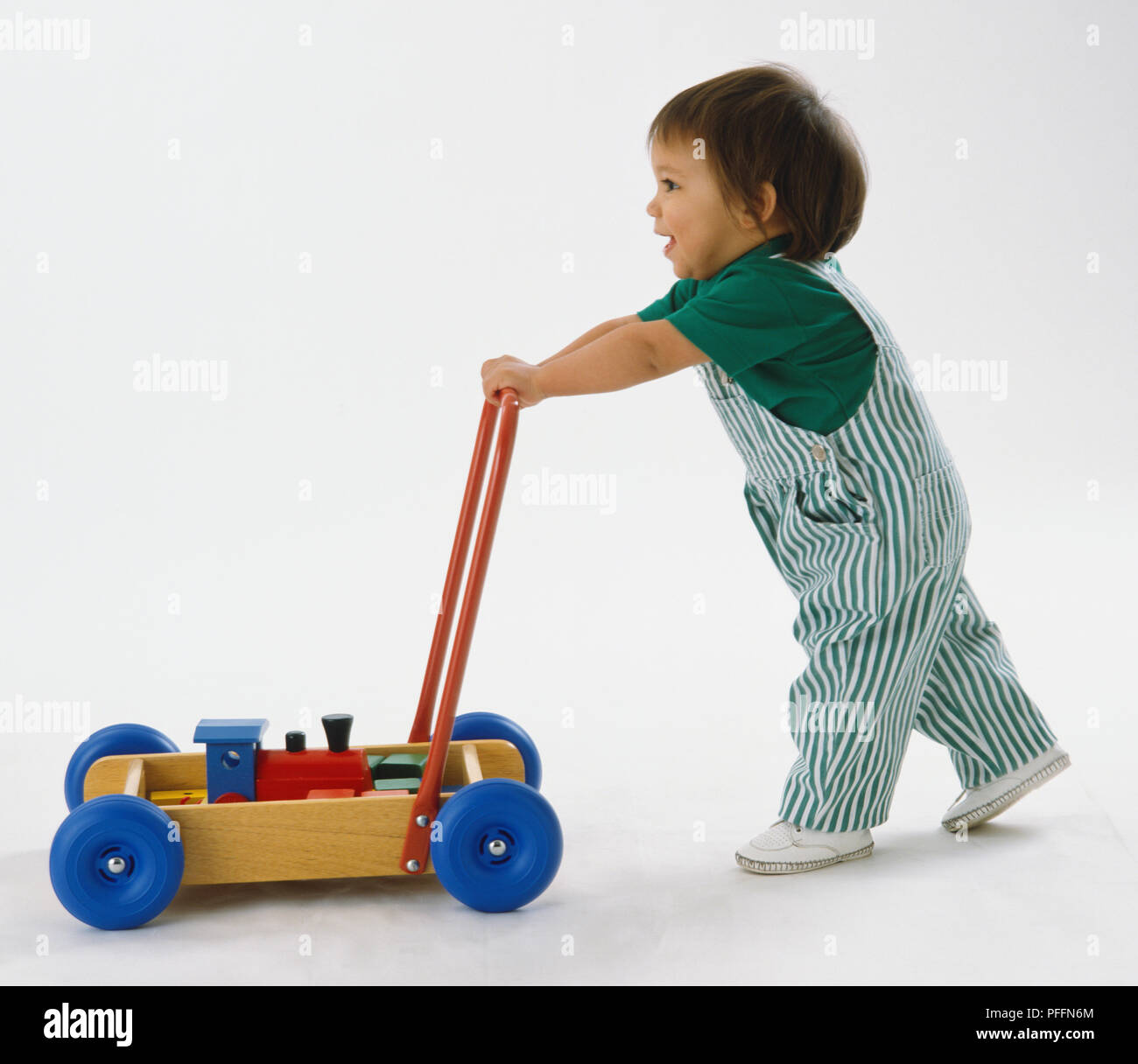 Toddler wearing striped dungarees, walking, pushing pushcart along ...