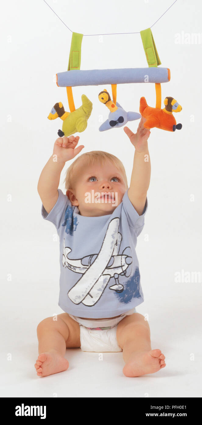 Baby boy with blonde hair, wearing nappy, sitting on floor, reaching up ...