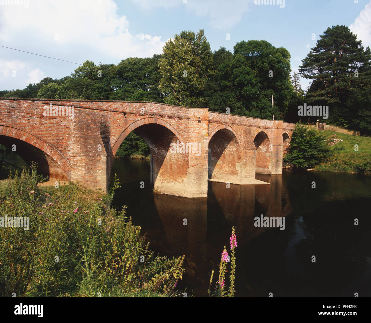 Dark river with a red brick bridge with four arches surrounded by trees ...