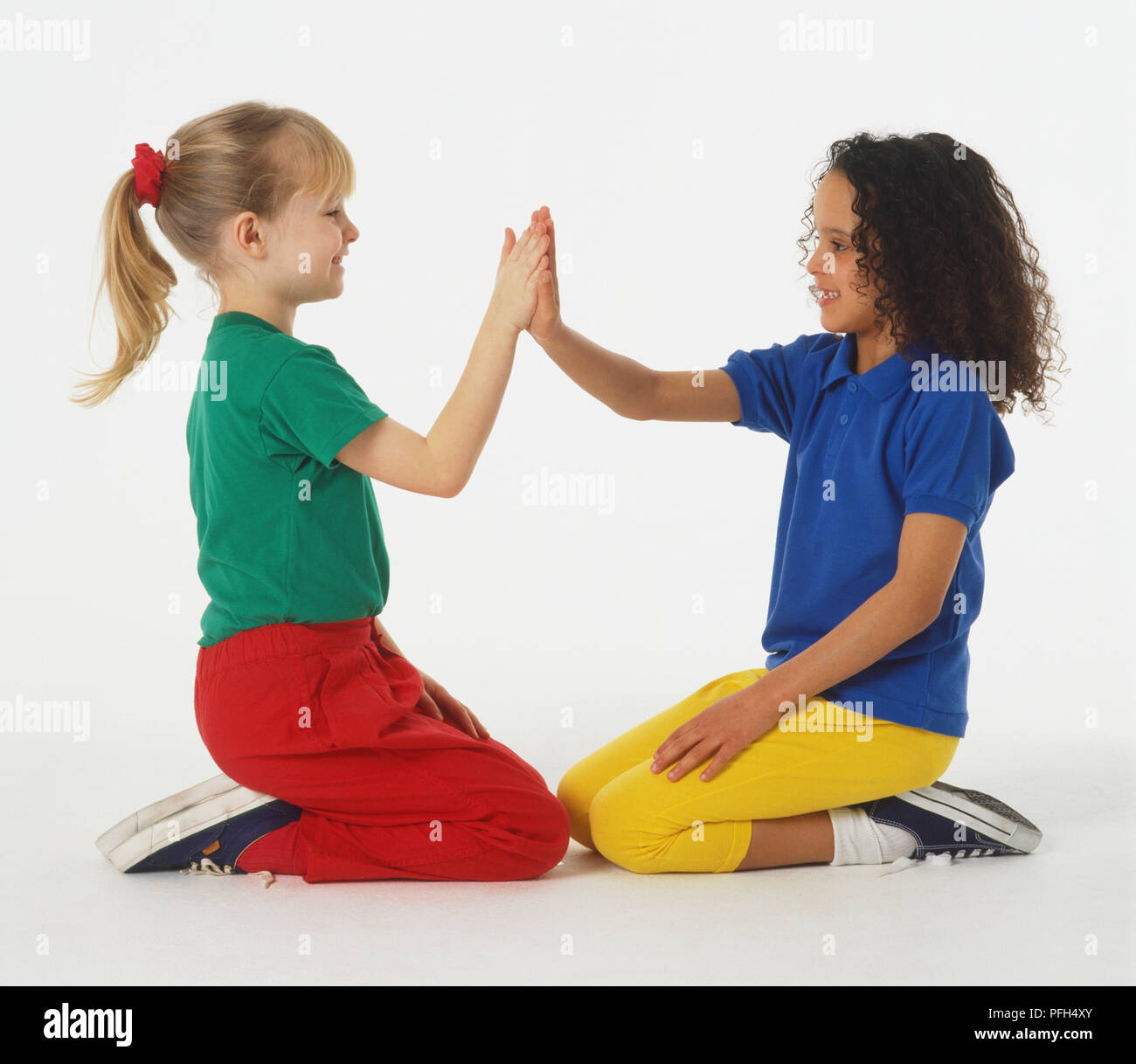 Two girls in colourful clothes sitting down on their knees and clapping ...