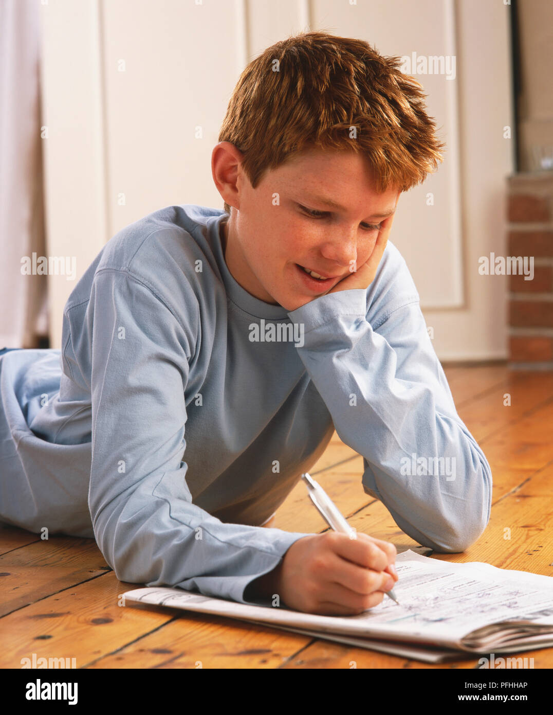 Teenage boy lying on wooden floor, noting down on a newspaper using a ...