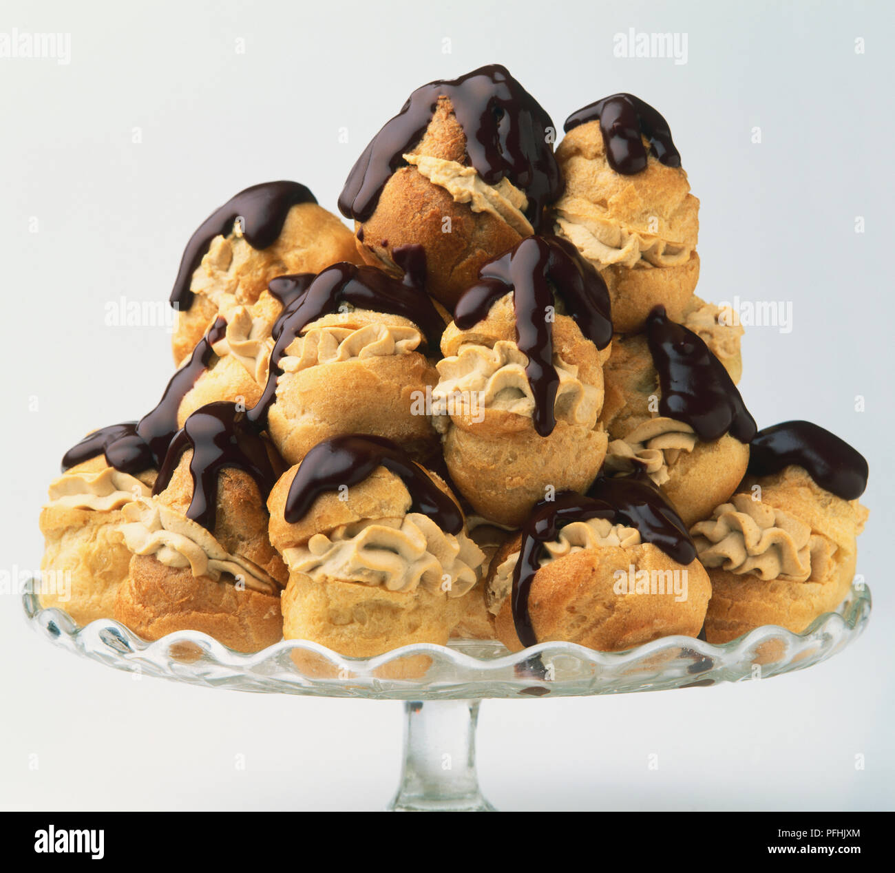 A mound of chocolate-topped profiteroles piled on glass cake stand ...