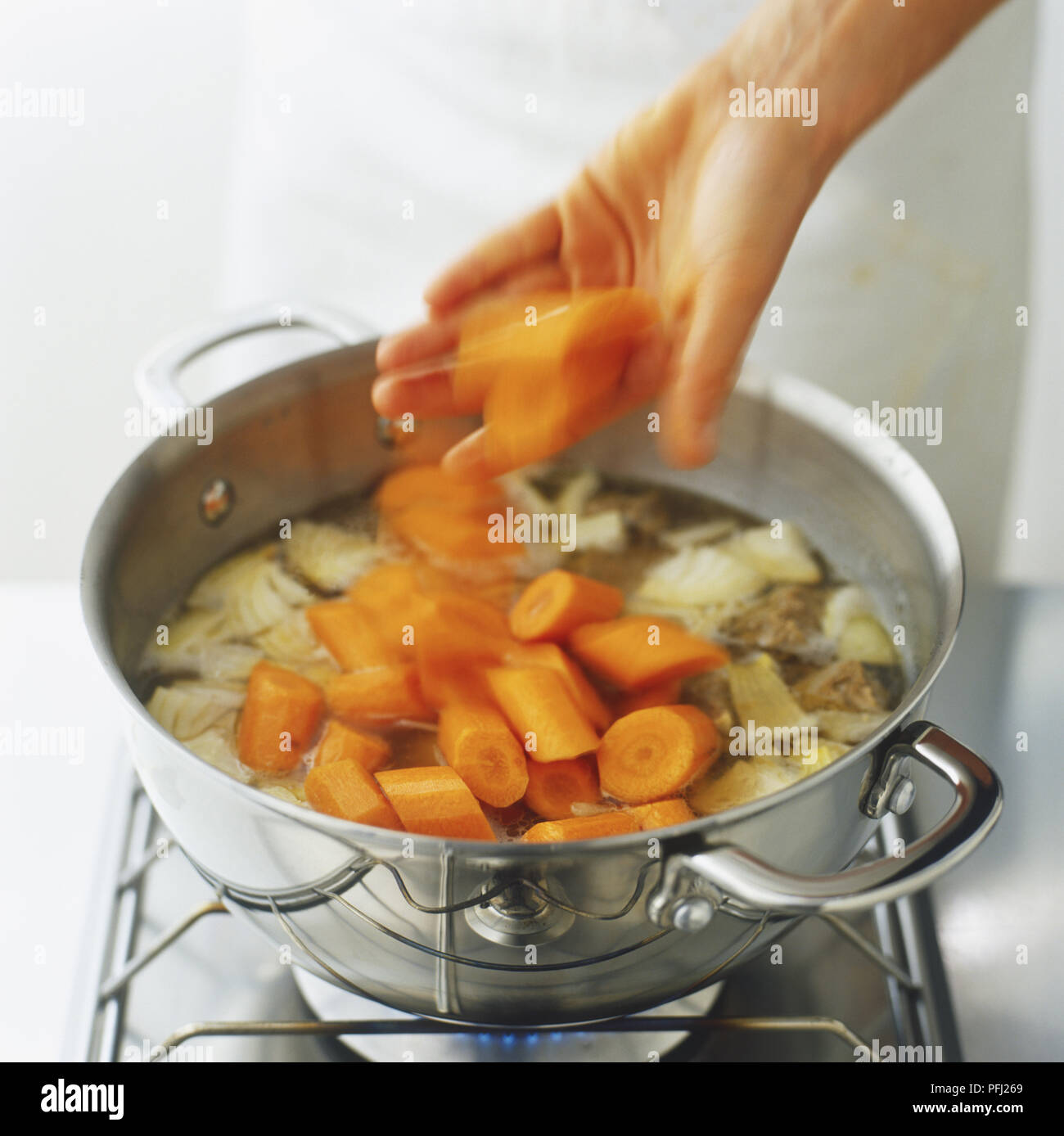 Adding carrots to a clear soup cooking in a pan on a gas stove, blurred ...