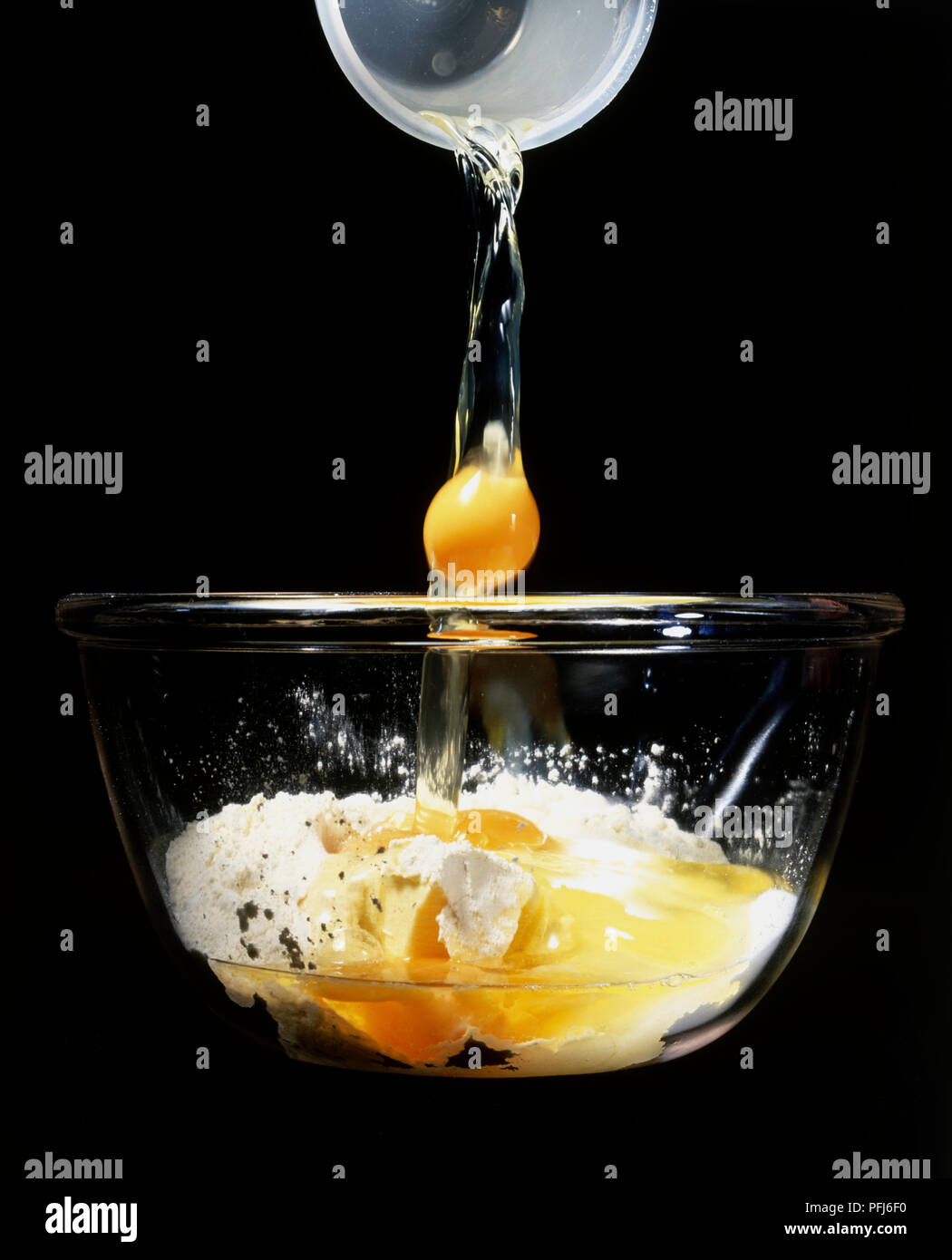 Raw egg being poured over sugar and flour in a glass mixing bowl, close ...