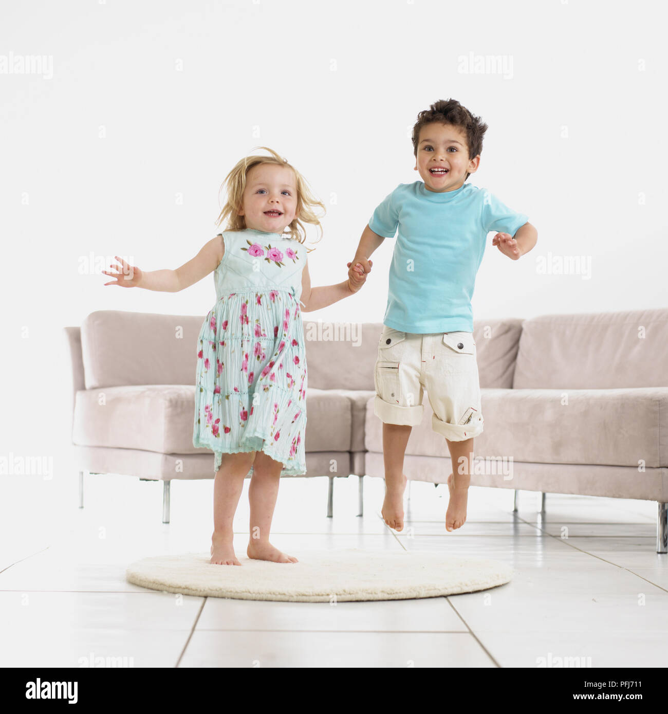 Girl and boy jumping up from a rug in living room, sofa in background ...