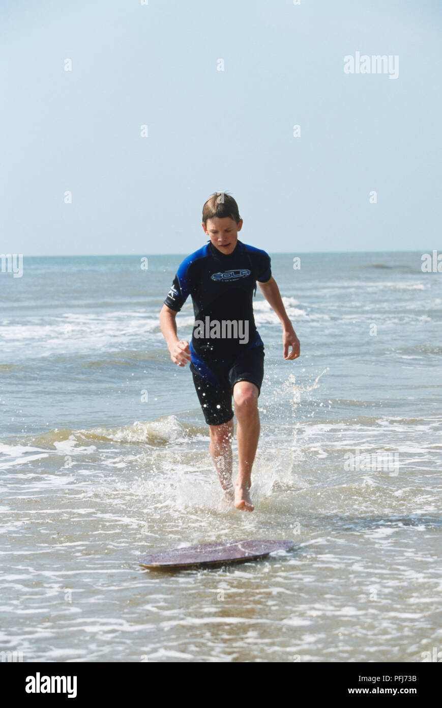 Boy wearing wetsuit running out of the sea in shallow surf towards ...