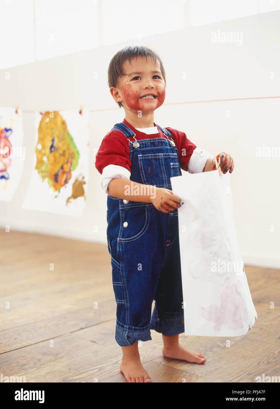 Smiling boy holding paper in hands, paintings hanging on line behind ...