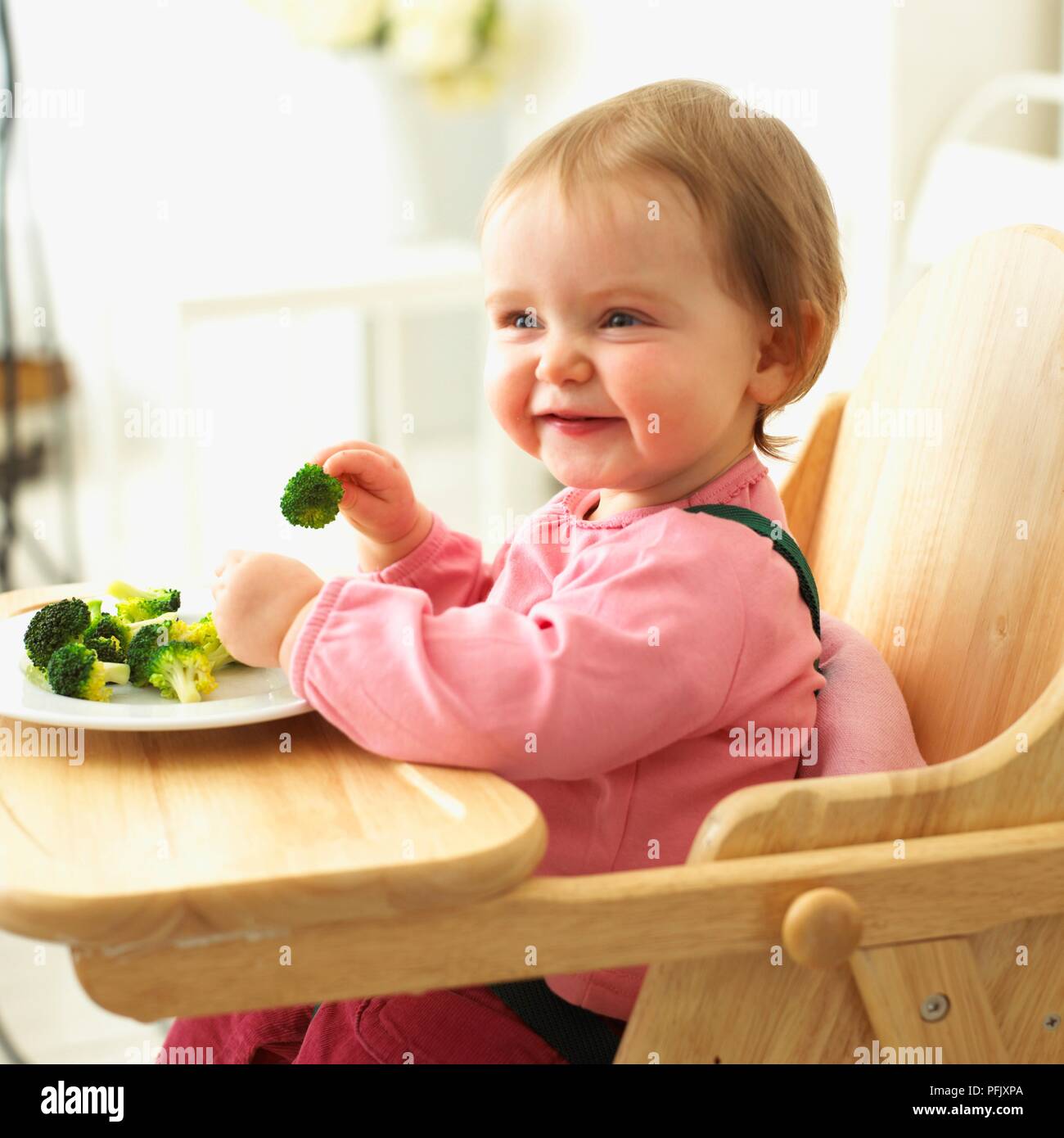 Baby girl sitting in wooden high chair eating broccoli, close-up Stock ...
