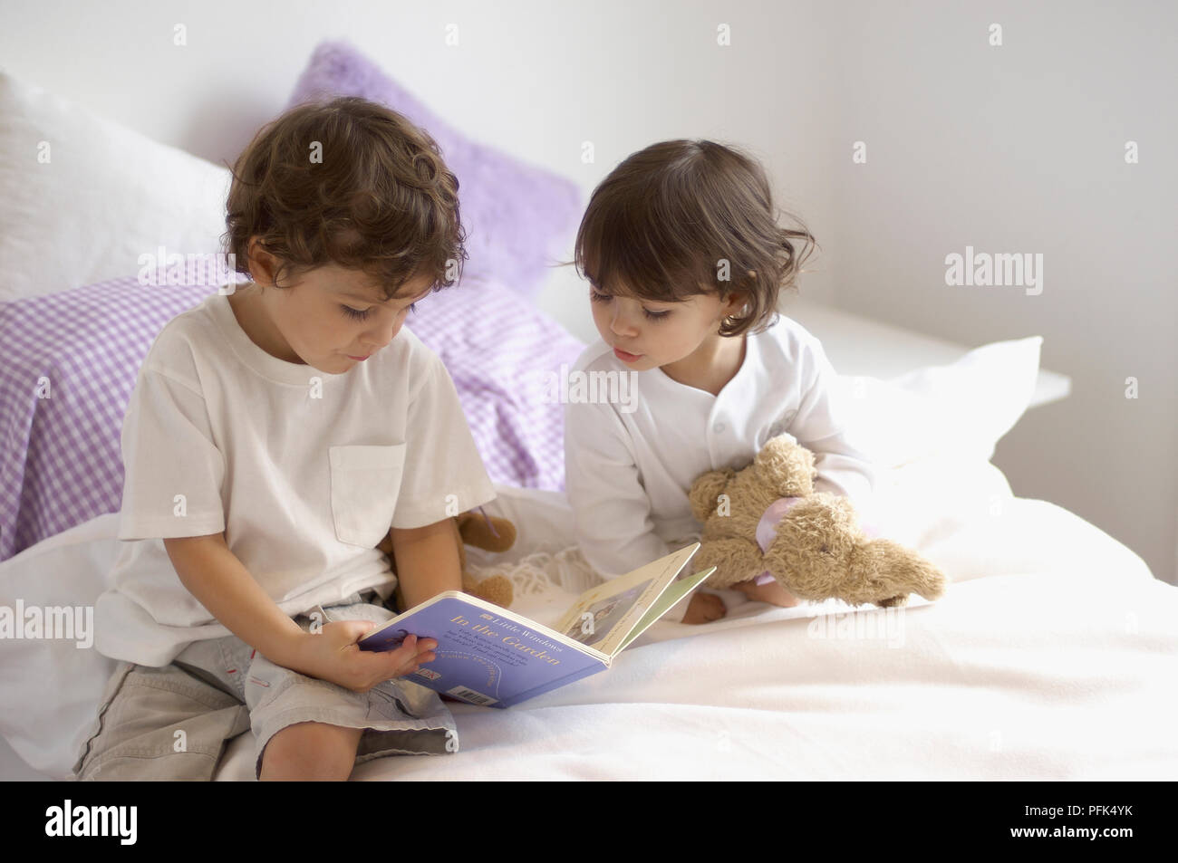 Young boy in pyjamas reading book to young girl lying in bed with teddy ...