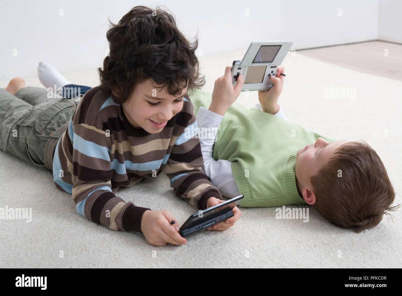 Two boys lying side by side on floor playing hand-held electronic games ...