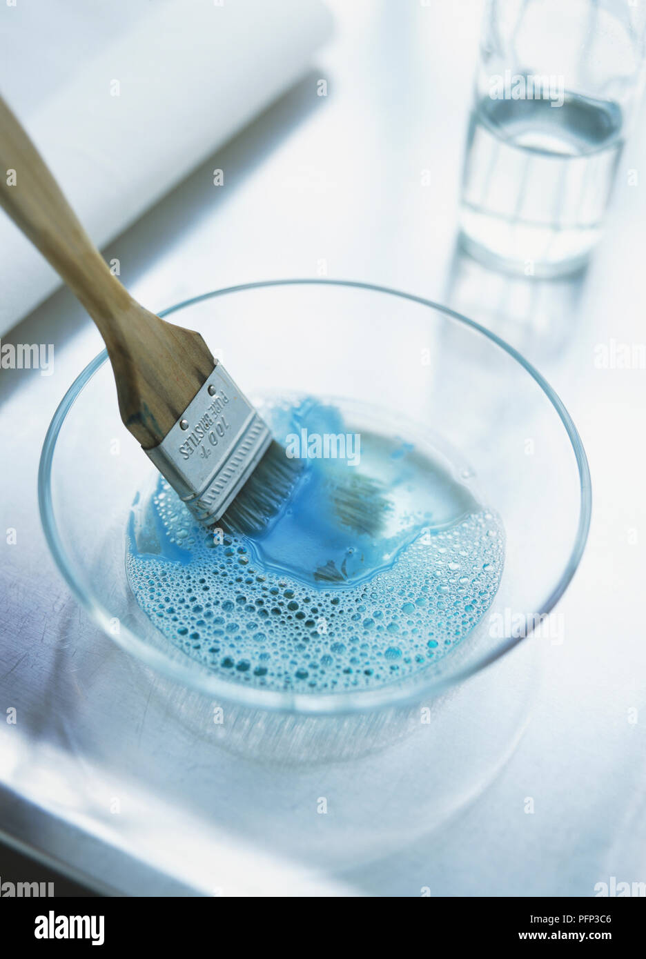 Paintbrush, with blue paint on bristles, soaking in glass bowl ...