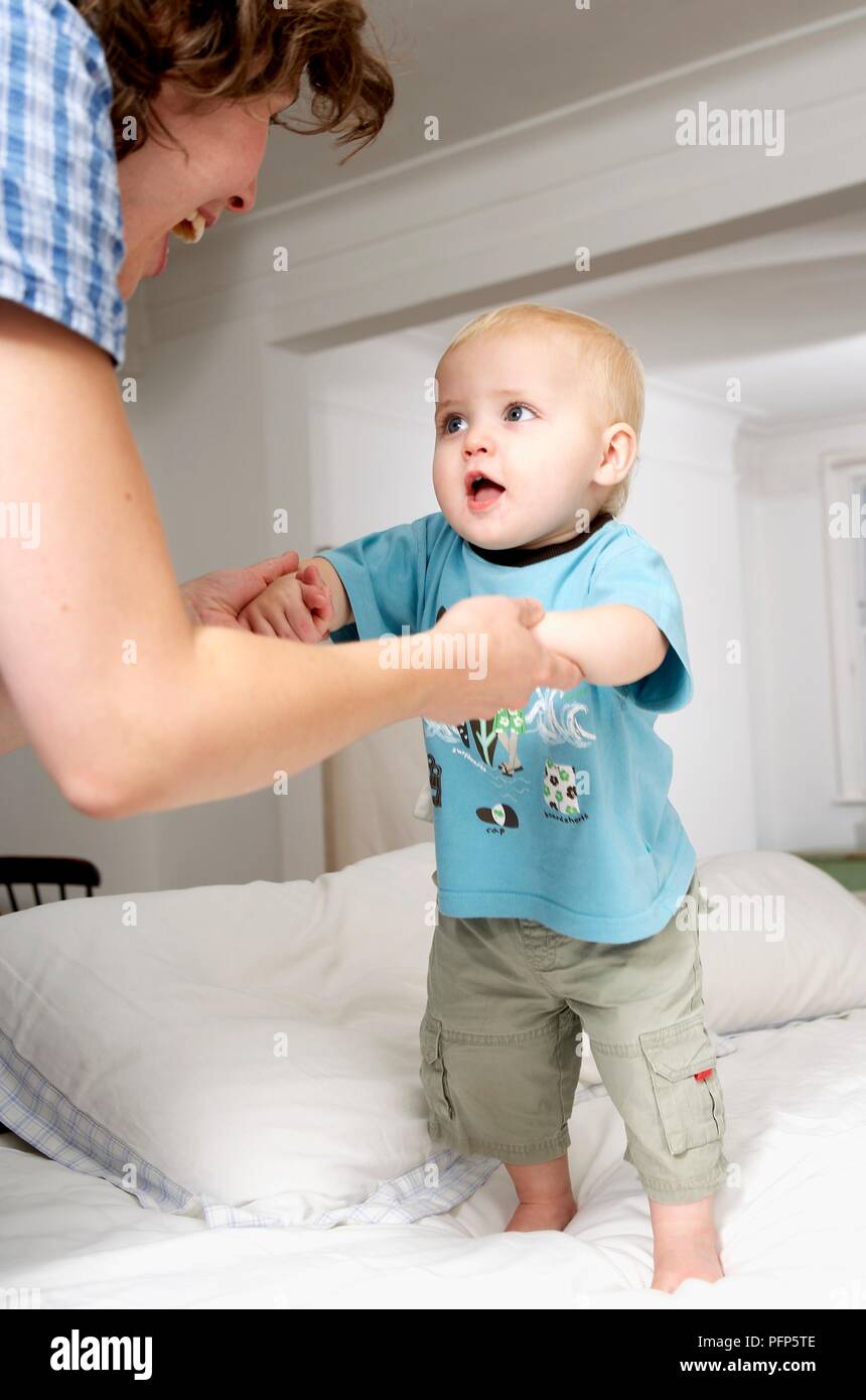 Baby boy standing up on bed supported by a woman's hands Stock Photo ...
