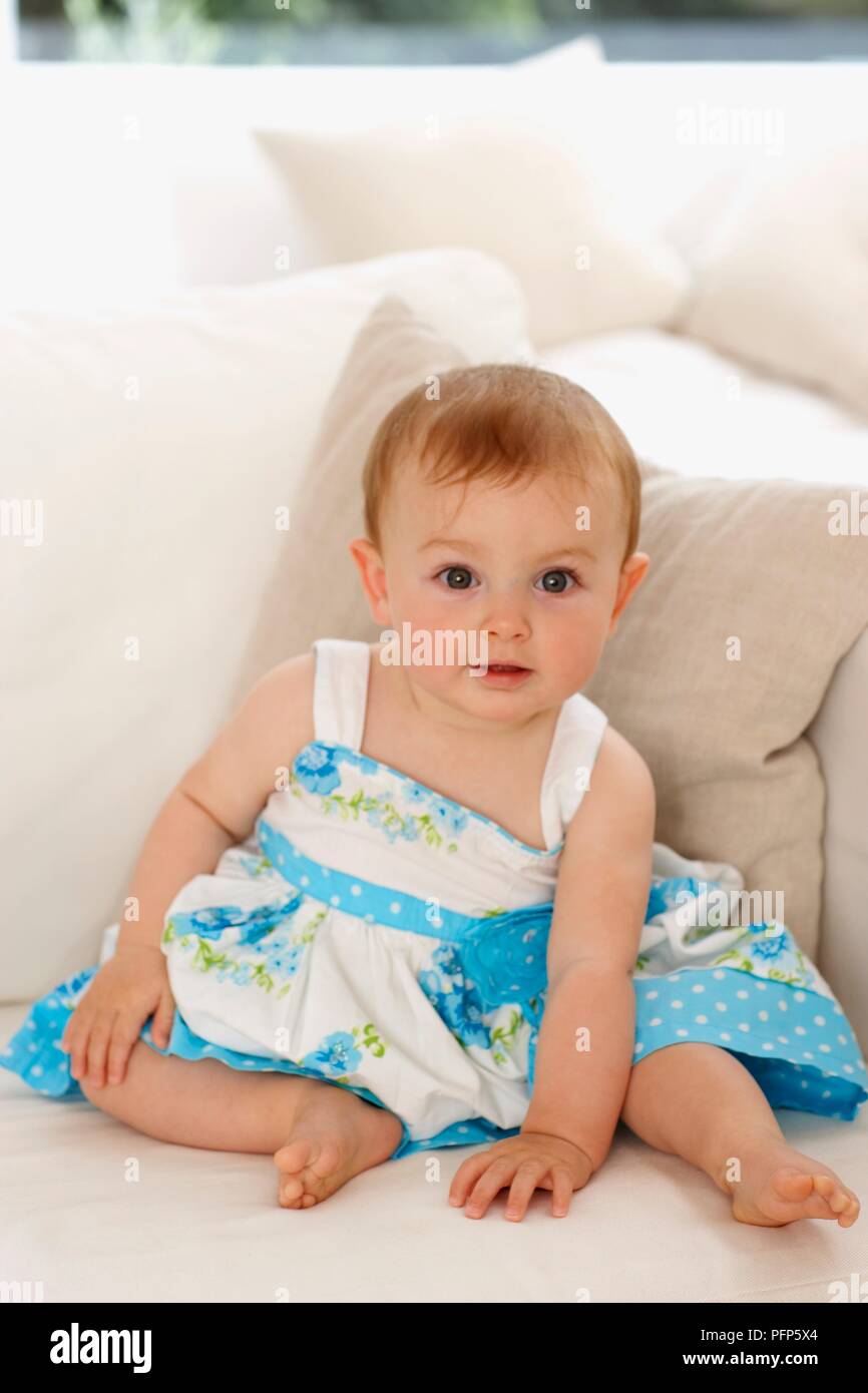 Baby girl in blue and white dress sitting on sofa, looking at camera ...