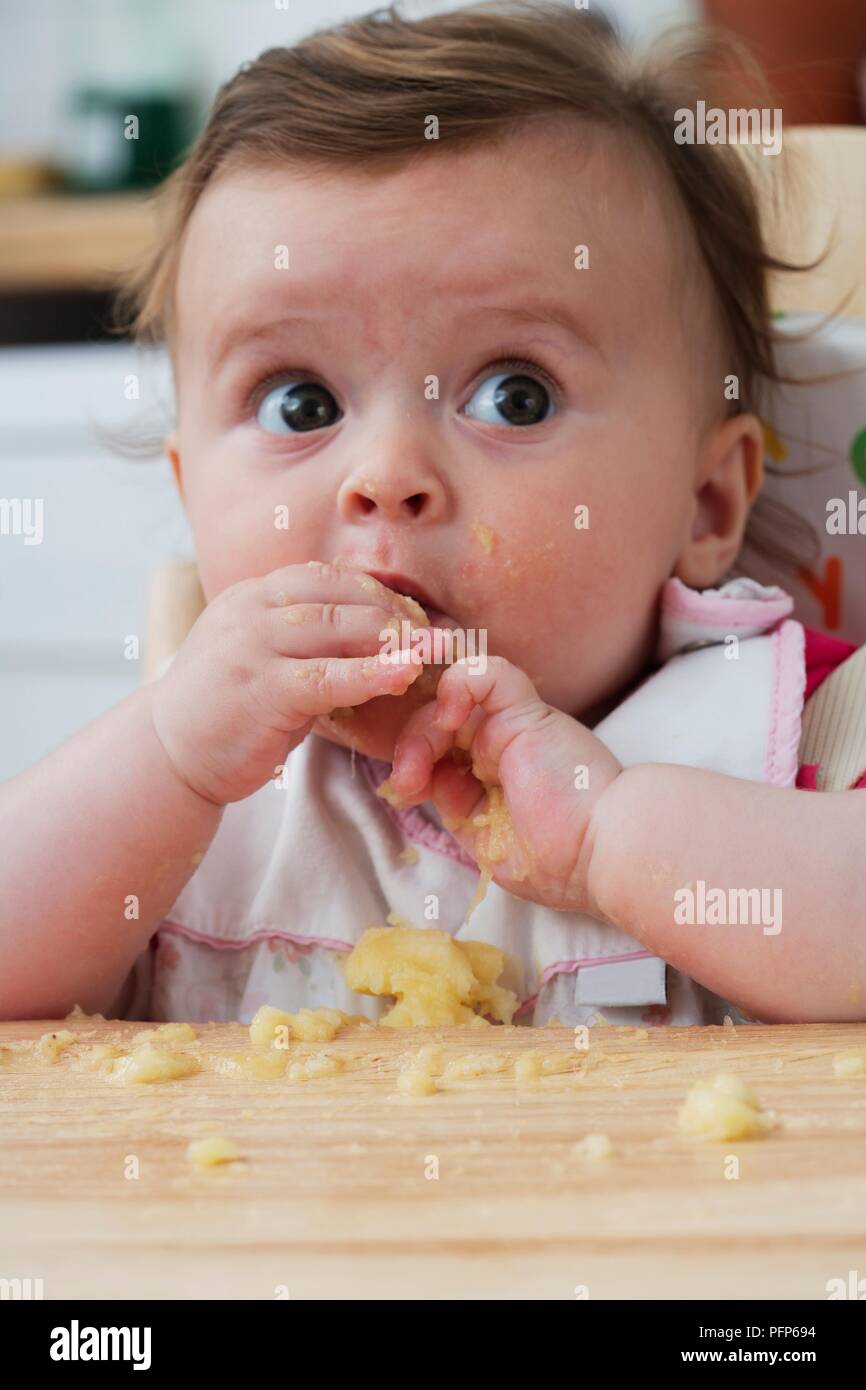 Head and shoulders of a baby girl eating a boiled potato, looking at ...