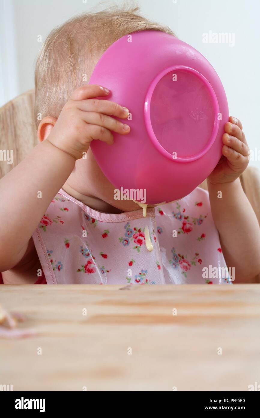 Baby girl in high chair holding pink plastic bowl over her face Stock Photo Alamy