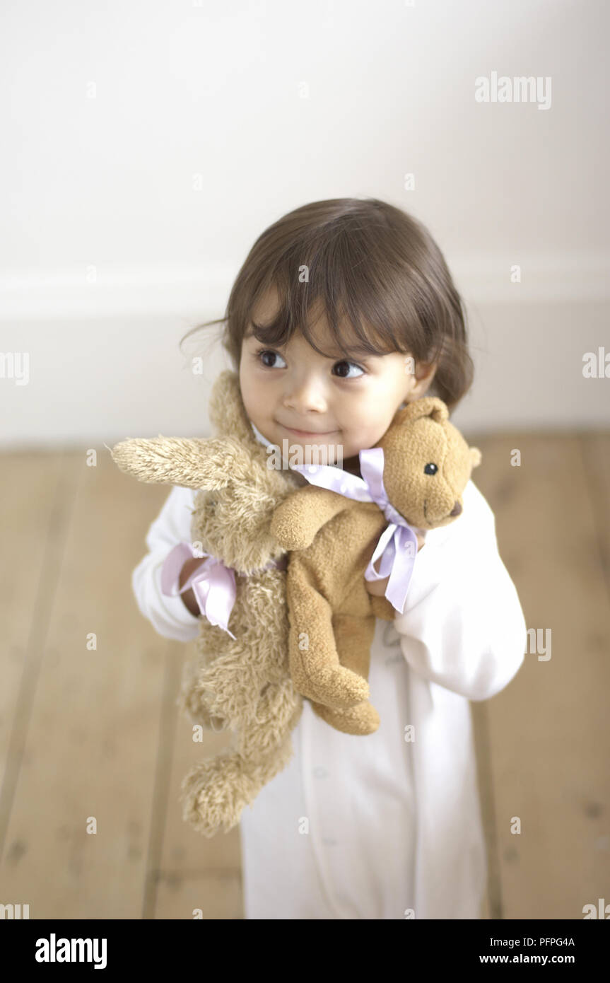 Young girl in pyjamas, standing, cuddling teddy bear and toy rabbit ...