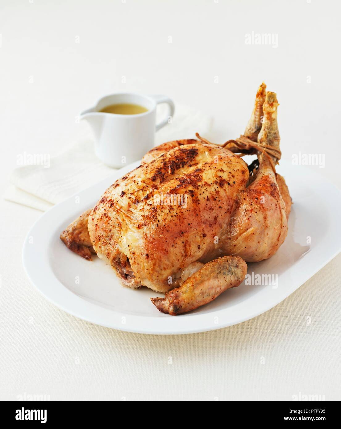 Stuffed roast chicken with legs tied together and jug of stock in ...