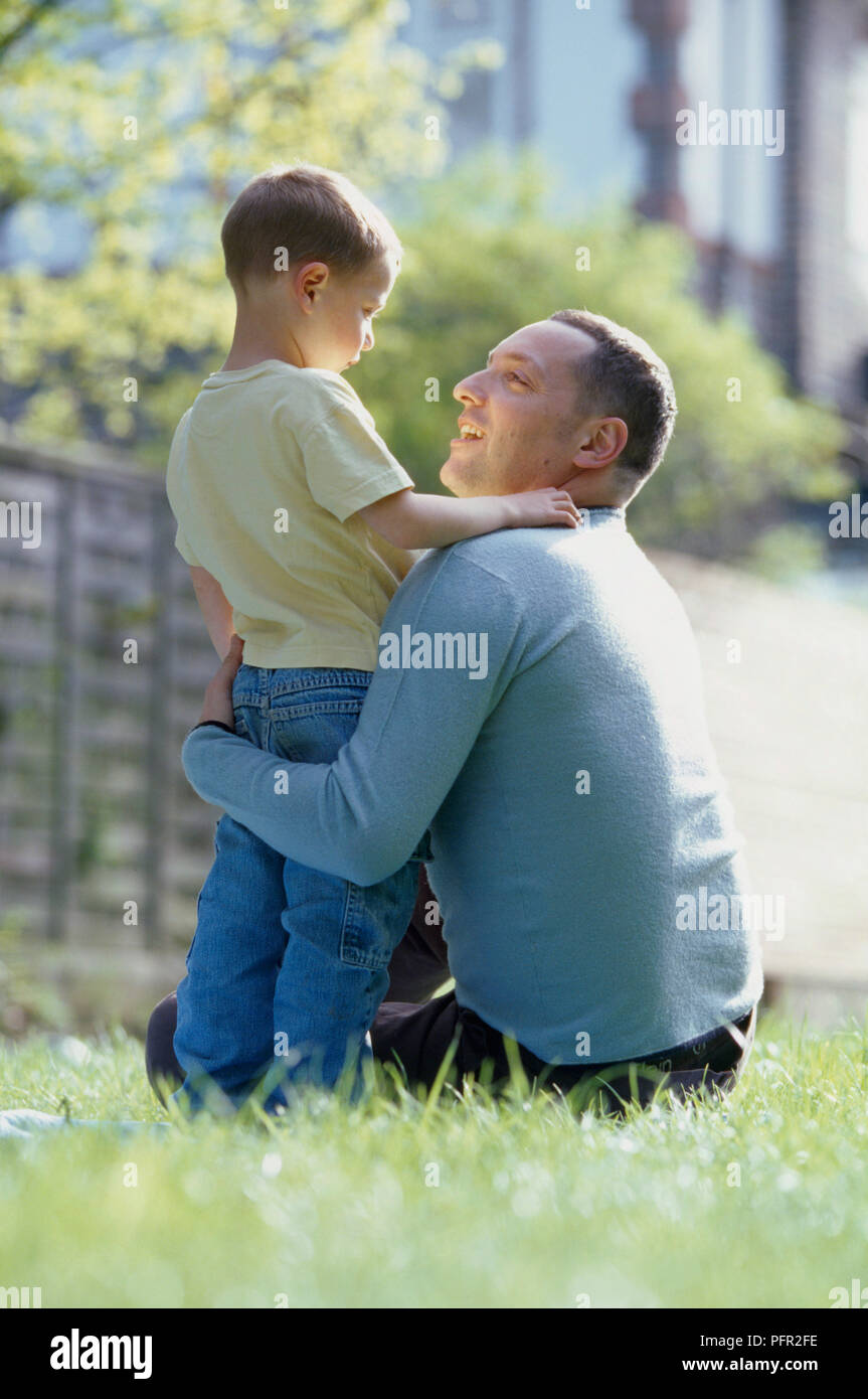 Man sitting down on grass, hugging boy around waist, looking at each ...