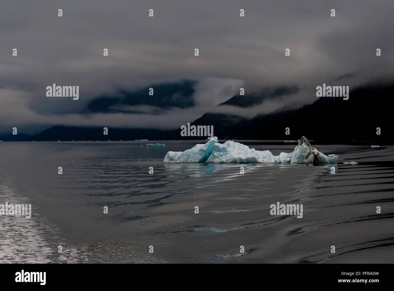 Alaska blue iceberg with cloudy mountains behind and wake from ship in ...