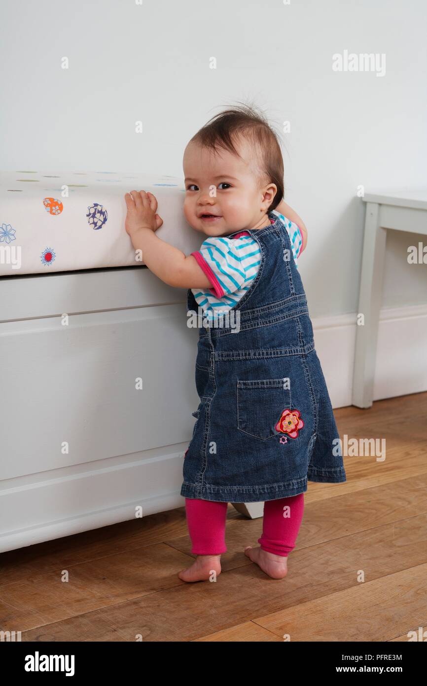 Baby girl standing next to a storage chest with her head turned to face ...