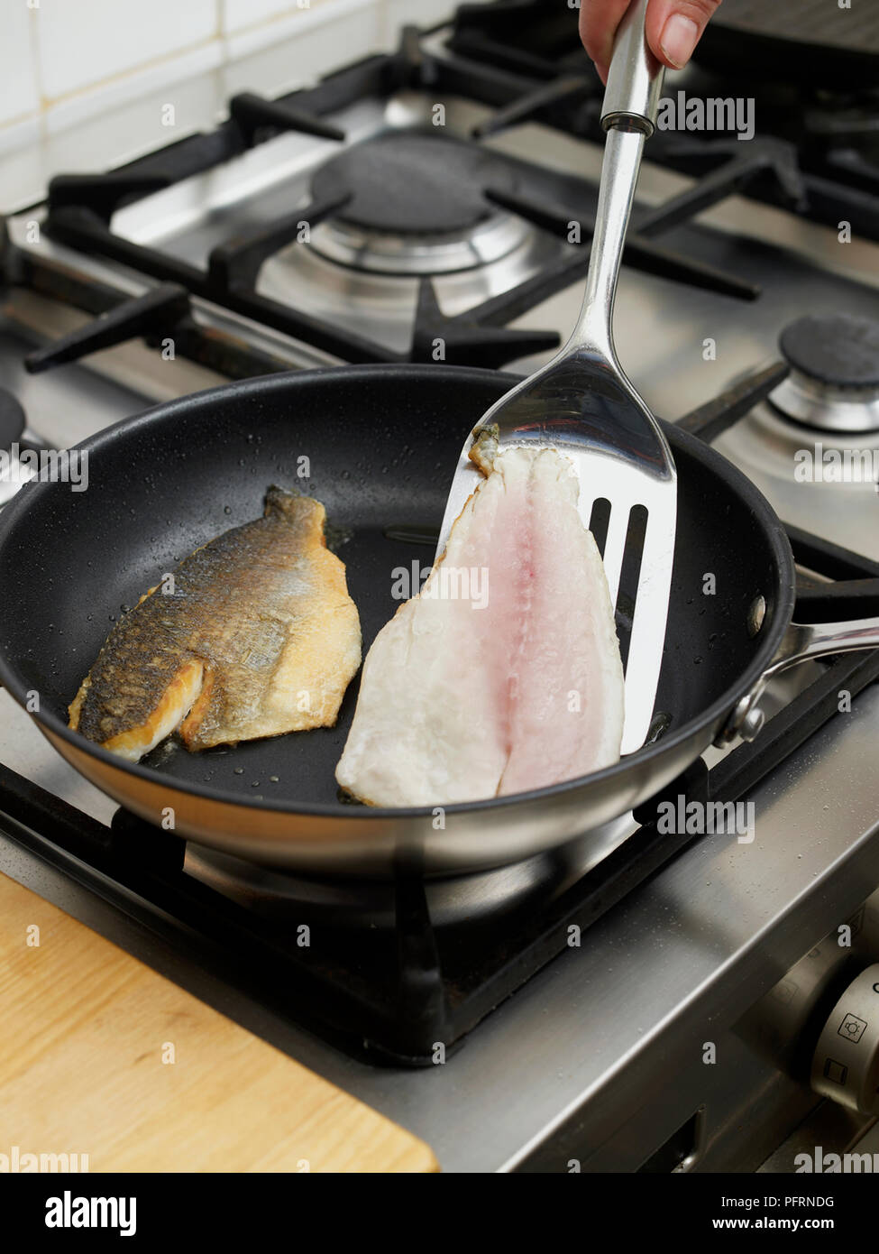 Using spatula to turn fish fillet in frying pan on gas hob, close-up ...