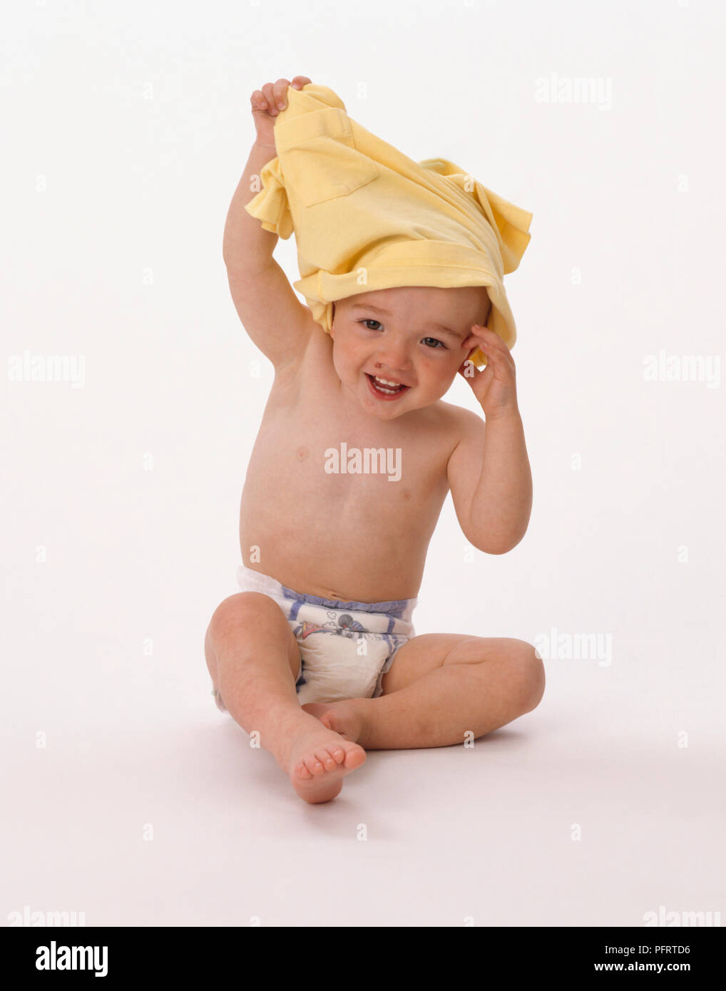 Smiling toddler sitting on floor wearing nappy and pulling yellow top ...