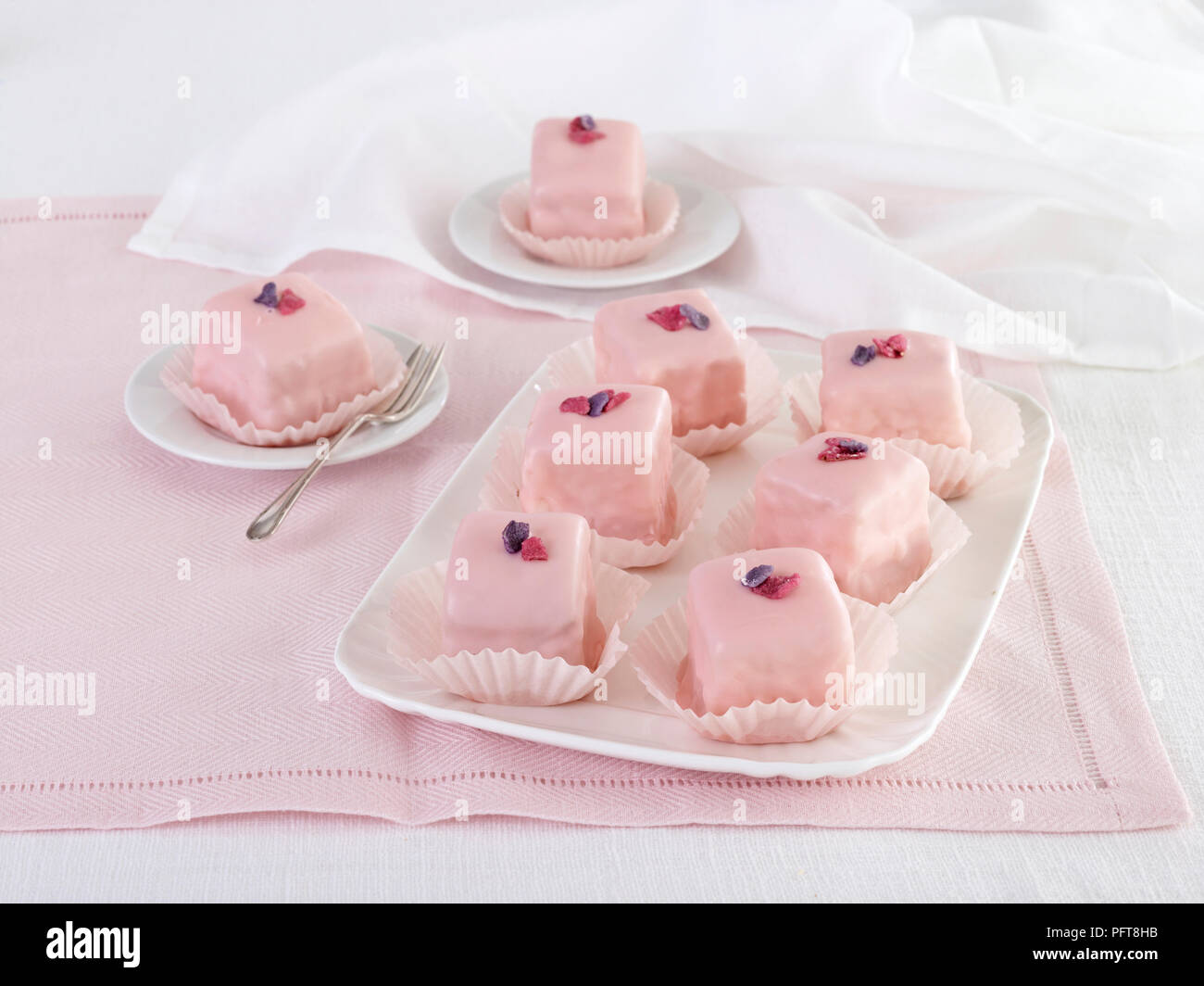 Pink icing fondant fancies on platter and plate with dessert fork and ...