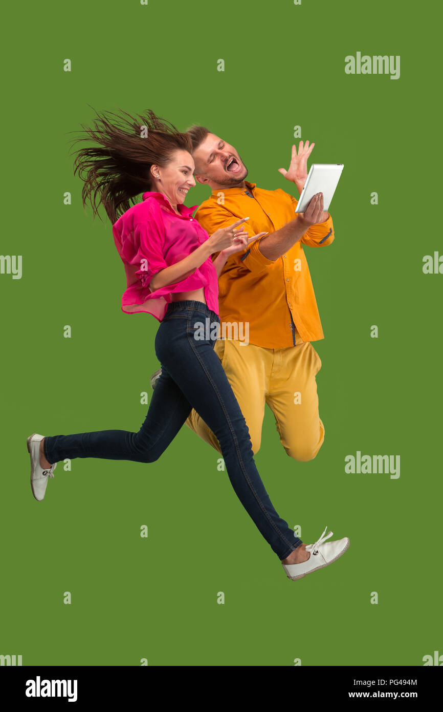 Jump of young couple over red studio background using laptop or tablet ...