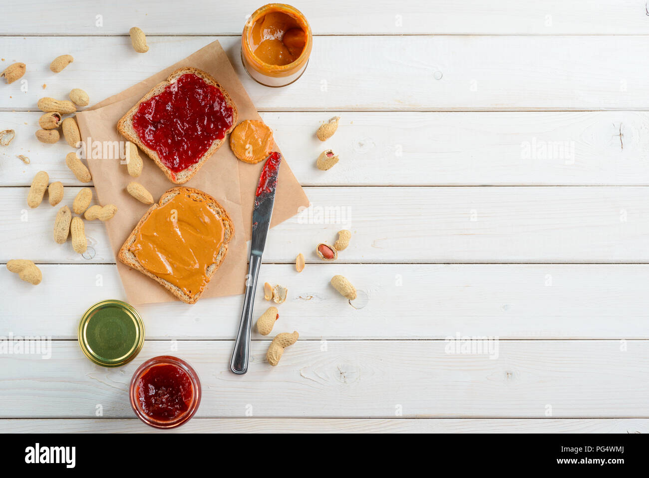 Peanuts, jam jar, sandwich and knife on baking paper. Light wooden ...
