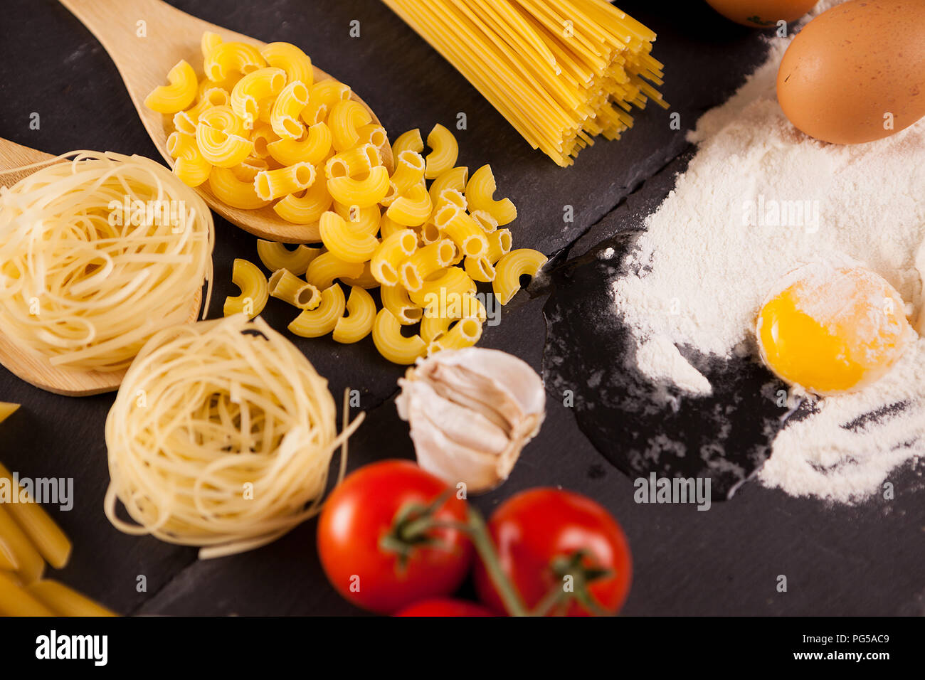Egg in flour next to different types of uncooked pasta on dark ...