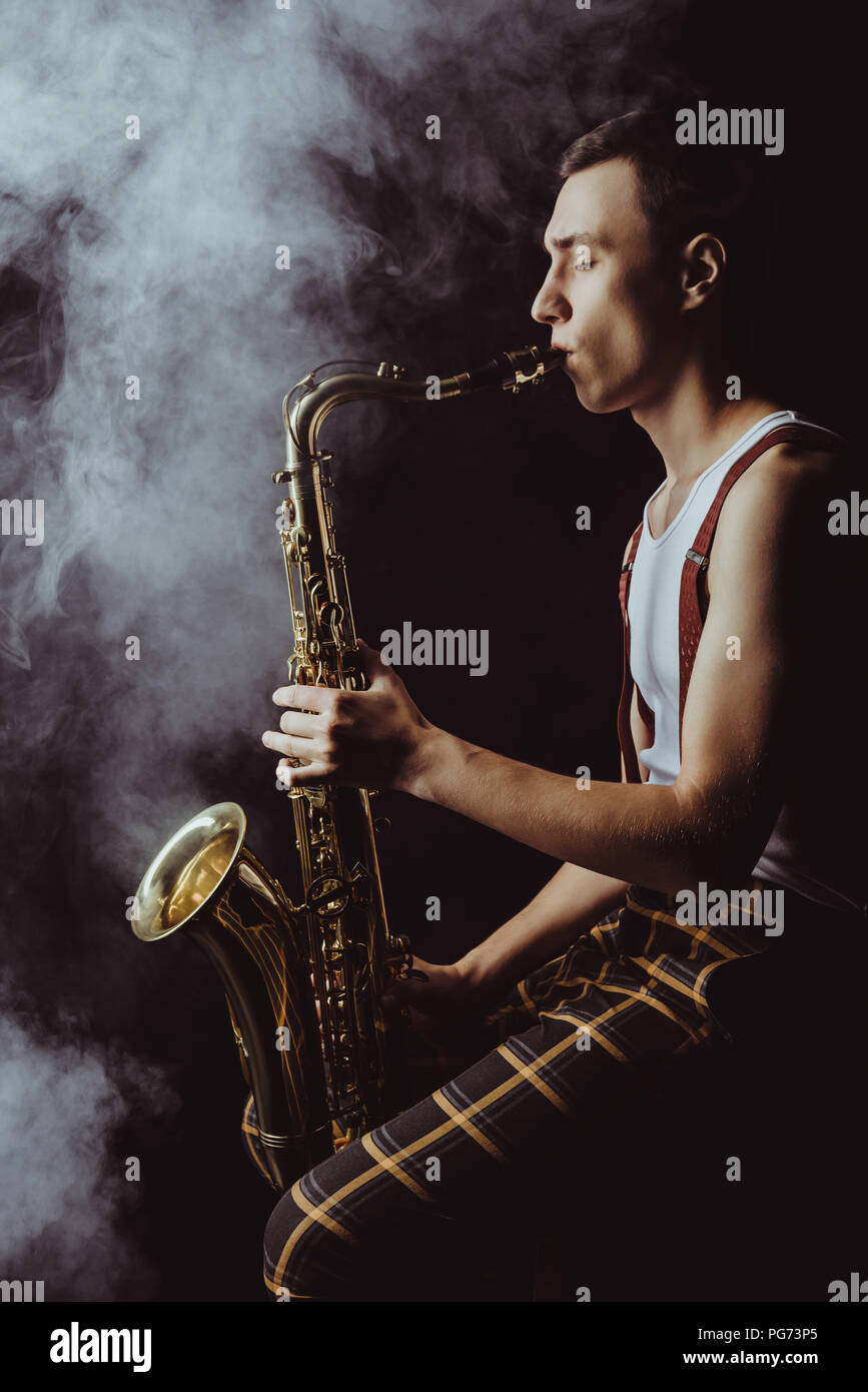 side view of stylish young jazzman sitting and playing saxophone in ...