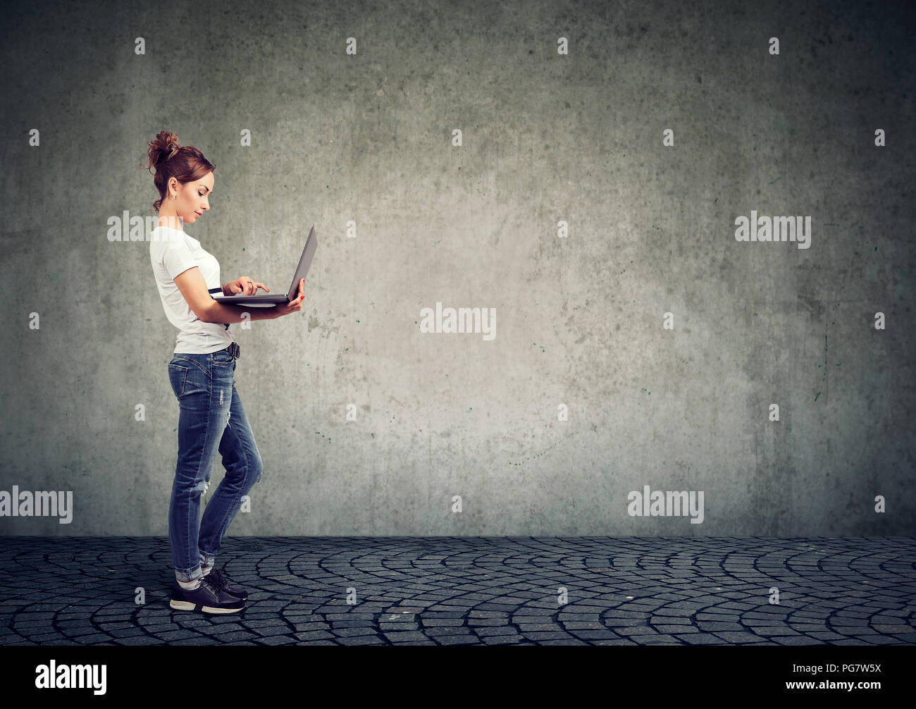 Full length portrait of a casual female student standing using laptop ...
