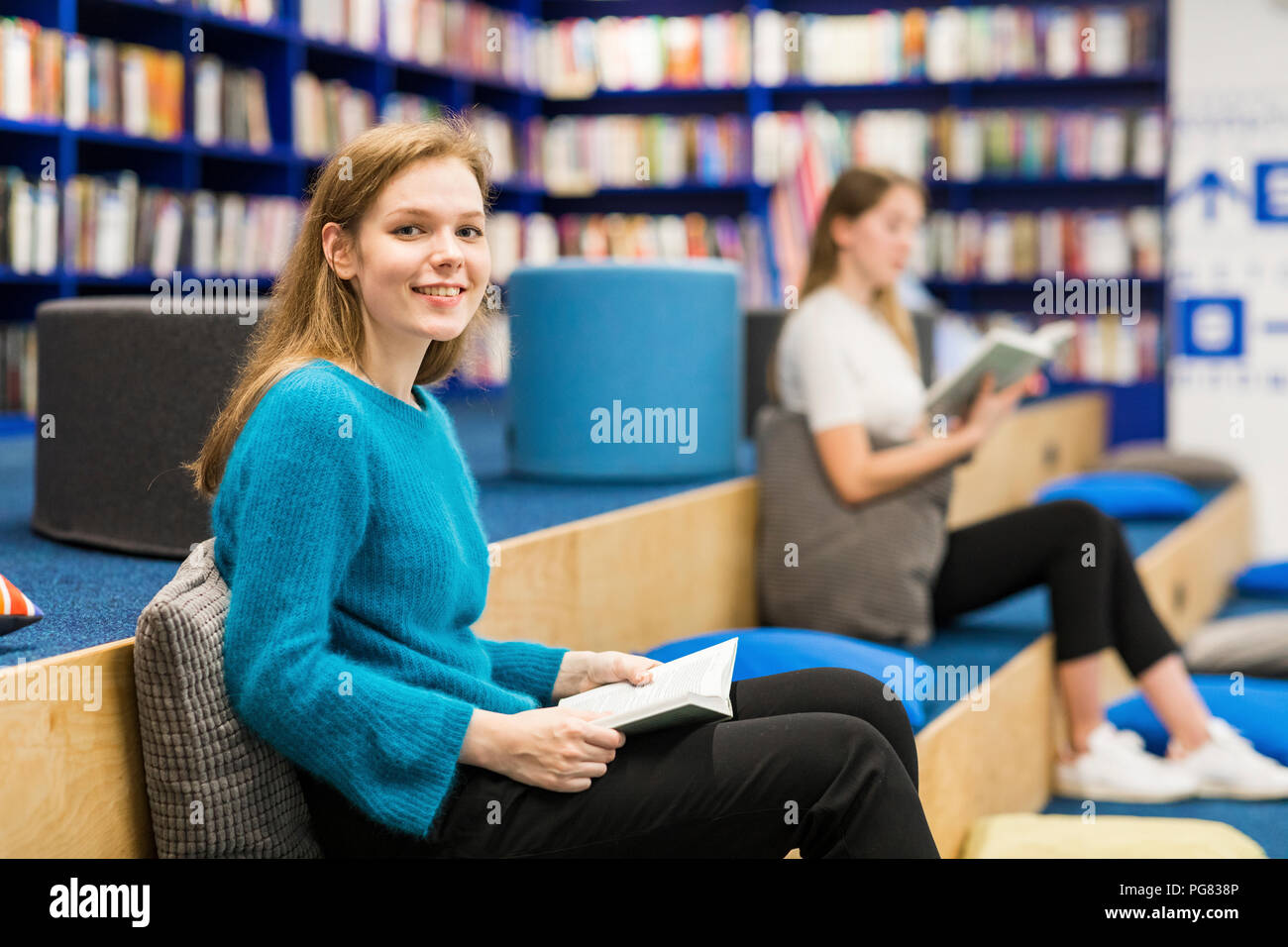 Portrait of smiling teenage girl sitting in public library reading book ...
