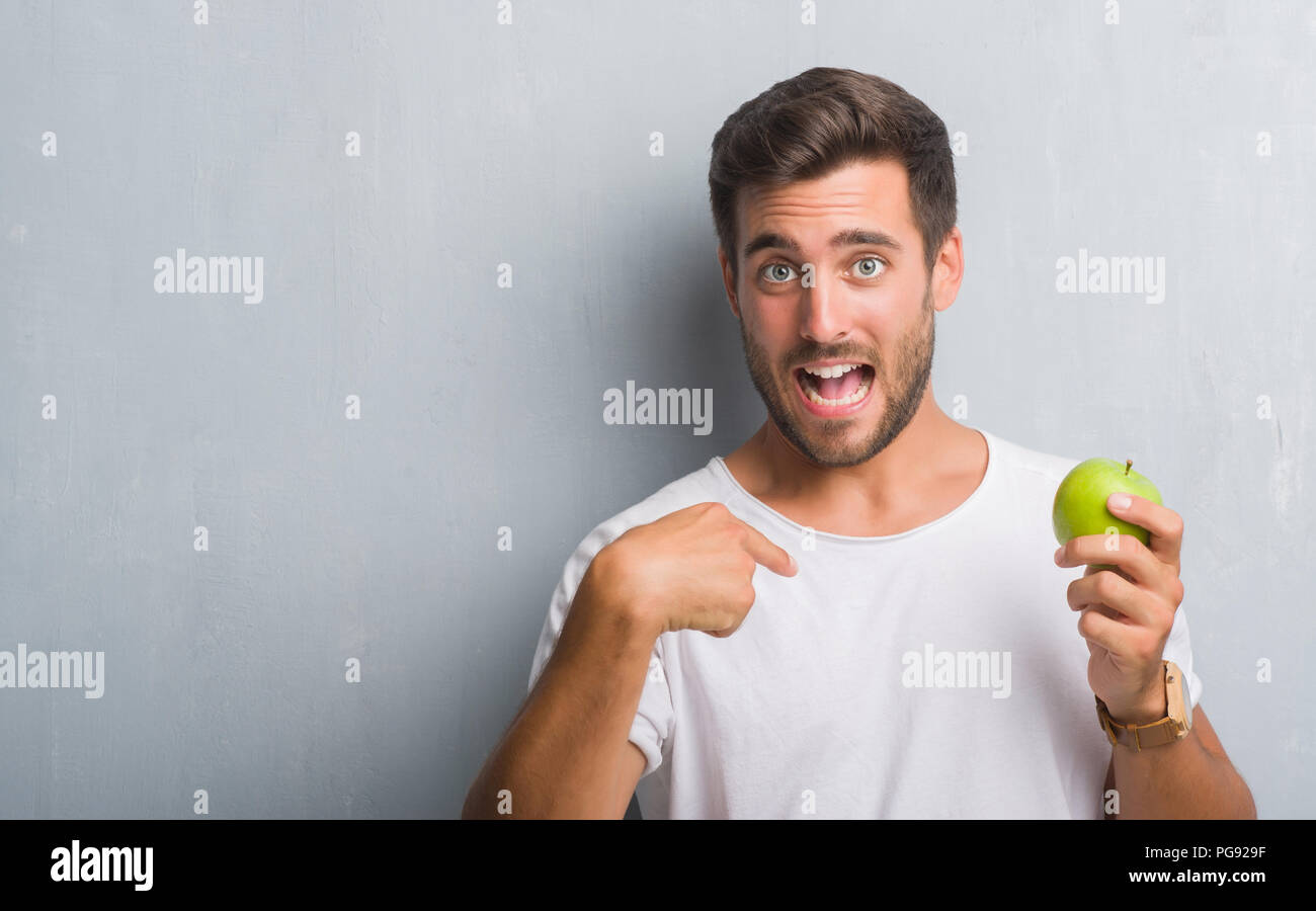 Handsome young man over grey grunge wall eating green apple with ...