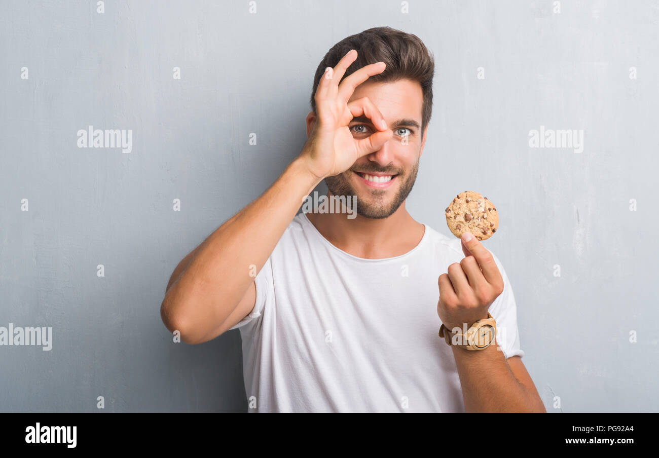 Handsome young man over grey grunge wall eating chocolate chip cooky ...