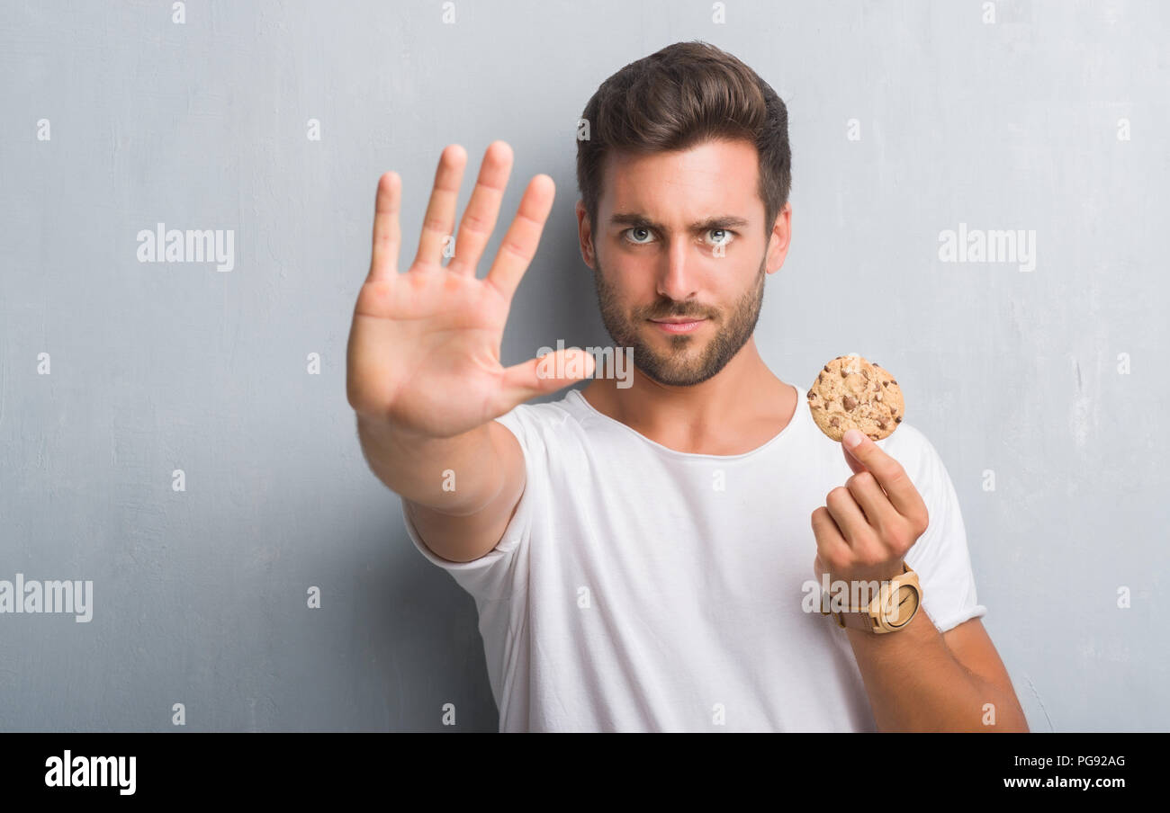 Handsome young man over grey grunge wall eating chocolate chip cooky ...