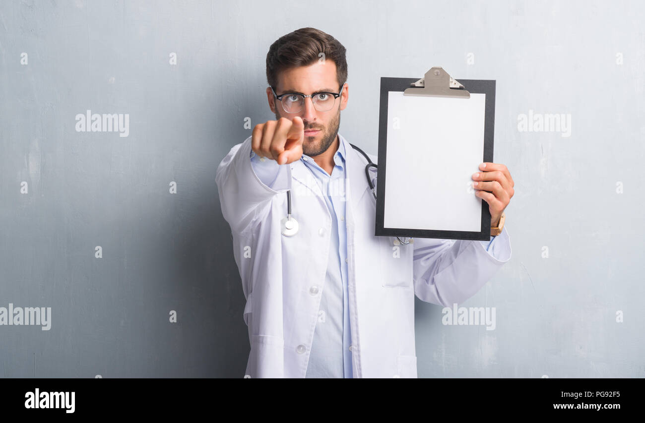 Handsome young doctor man over grey grunge wall holding clipboard ...