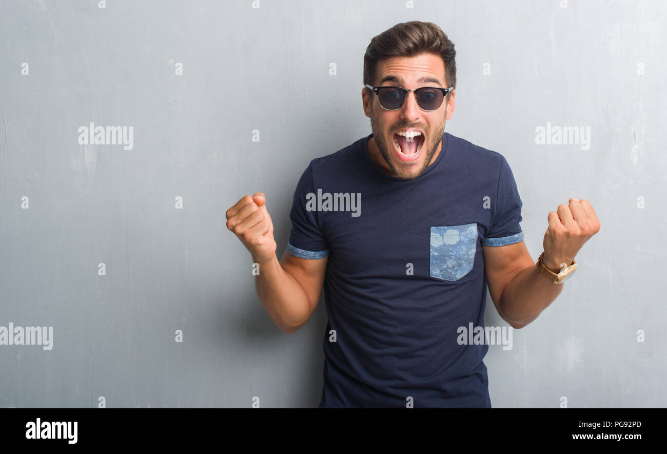 Handsome Young Man Over Grey Grunge Wall Wearing Sunglasses Celebrating